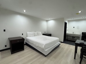 Quarto moderno com cama, mesas de cabeceira, escrivaninha e banheiro com espelho e pia.