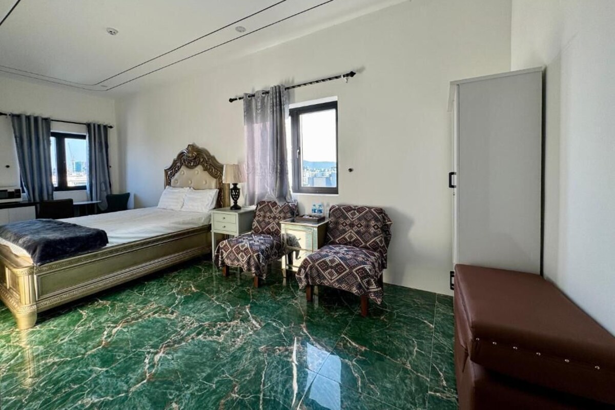 Quarto com cama, duas cadeiras, mesa de cabeceira, abajur e vista para a cidade.
