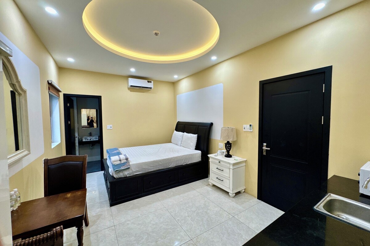 Quarto de hotel com cama, mesa de cabeceira, abajur e porta preta.