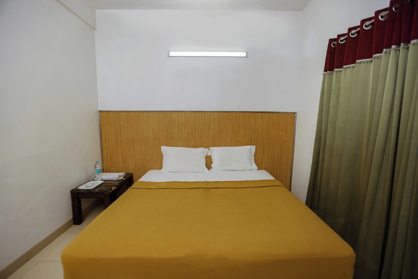 Quarto de hotel com cama, mesa de cabeceira, luminária, janela com cortinas e luminária fixada na parede.