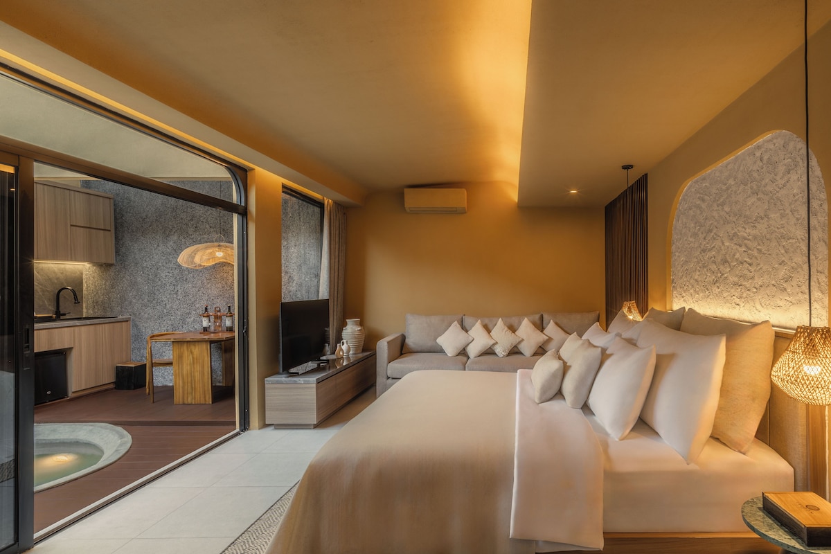 Quarto de hotel moderno com uma cama grande, um sofá, uma área de refeições e vista para a cozinha.