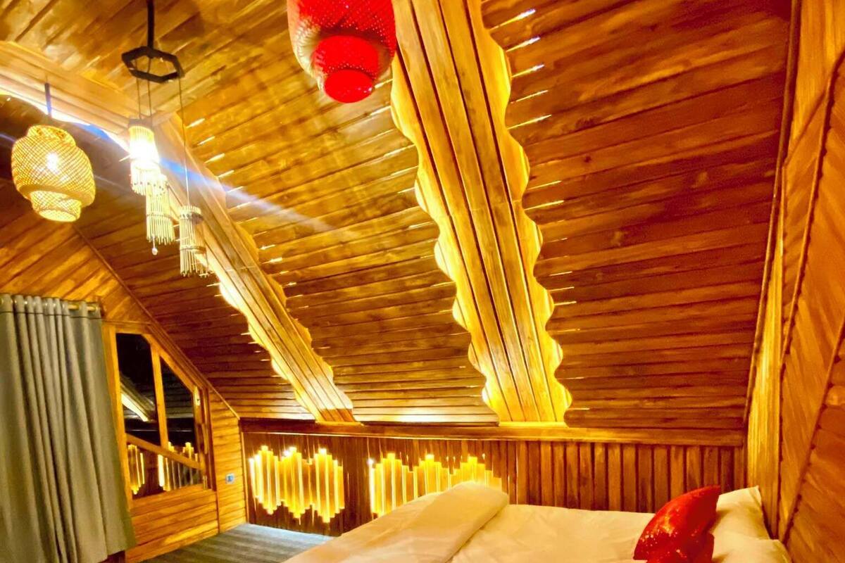 Quarto com teto inclinado, paredes de madeira, cama com roupa de cama branca e luminárias pendentes.