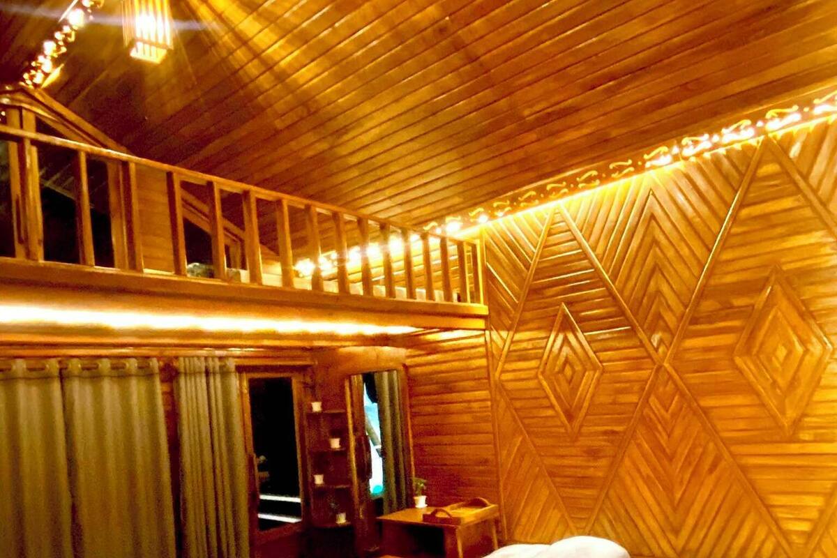 Quarto de hotel com teto de madeira, cama com roupa de cama branca, uma mesinha e uma cadeira.