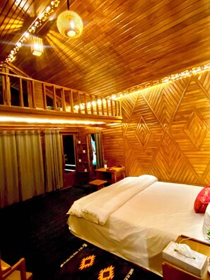 Quarto de hotel com teto de madeira, cama com roupa de cama branca, uma mesinha e uma cadeira.