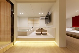 Quarto de hotel moderno com chão de madeira, uma pequena mesa com cadeiras, uma janela grande com persianas, uma TV fixada na parede e uma cama com roupa de cama branca.