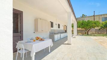Uma área de refeições ao ar livre com uma mesa posta para dois, uma cozinha ao fundo e vista para o jardim.
