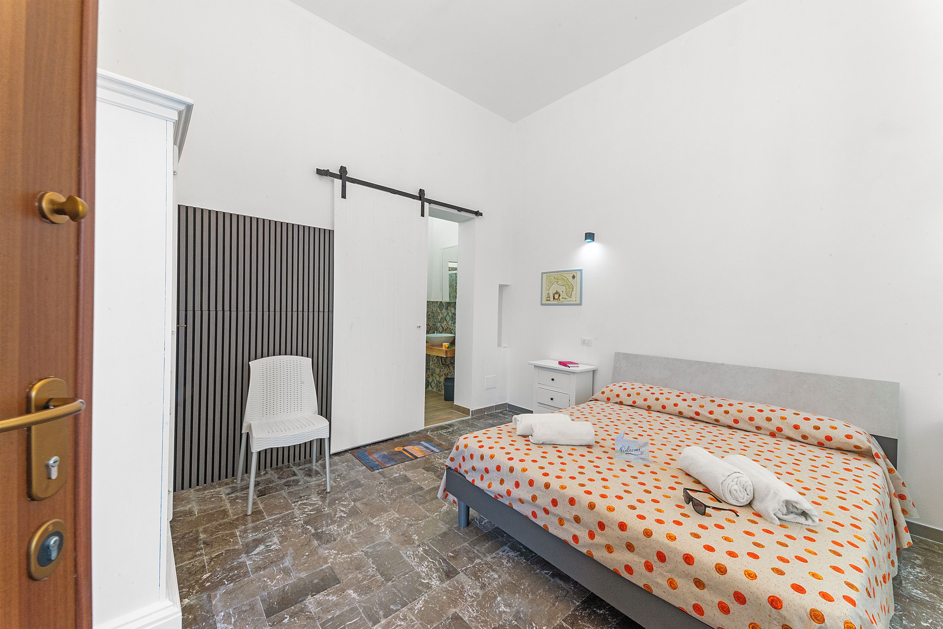 Um quarto com cama, uma cadeira, uma mesa pequena e uma porta.
