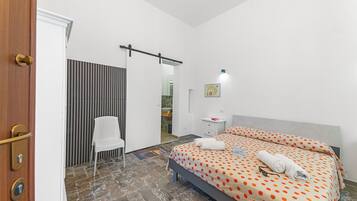 Um quarto com cama, uma cadeira, uma mesa pequena e uma porta.
