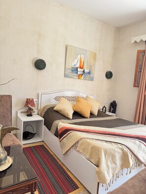Quarto com cama, mesa de cabeceira, luminária, almofadas decorativas, um quadro e um tapete estampado.