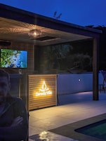 Uma área externa moderna com piscina, uma estrutura de madeira com um letreiro de neon e uma tela de televisão exibindo uma praia.