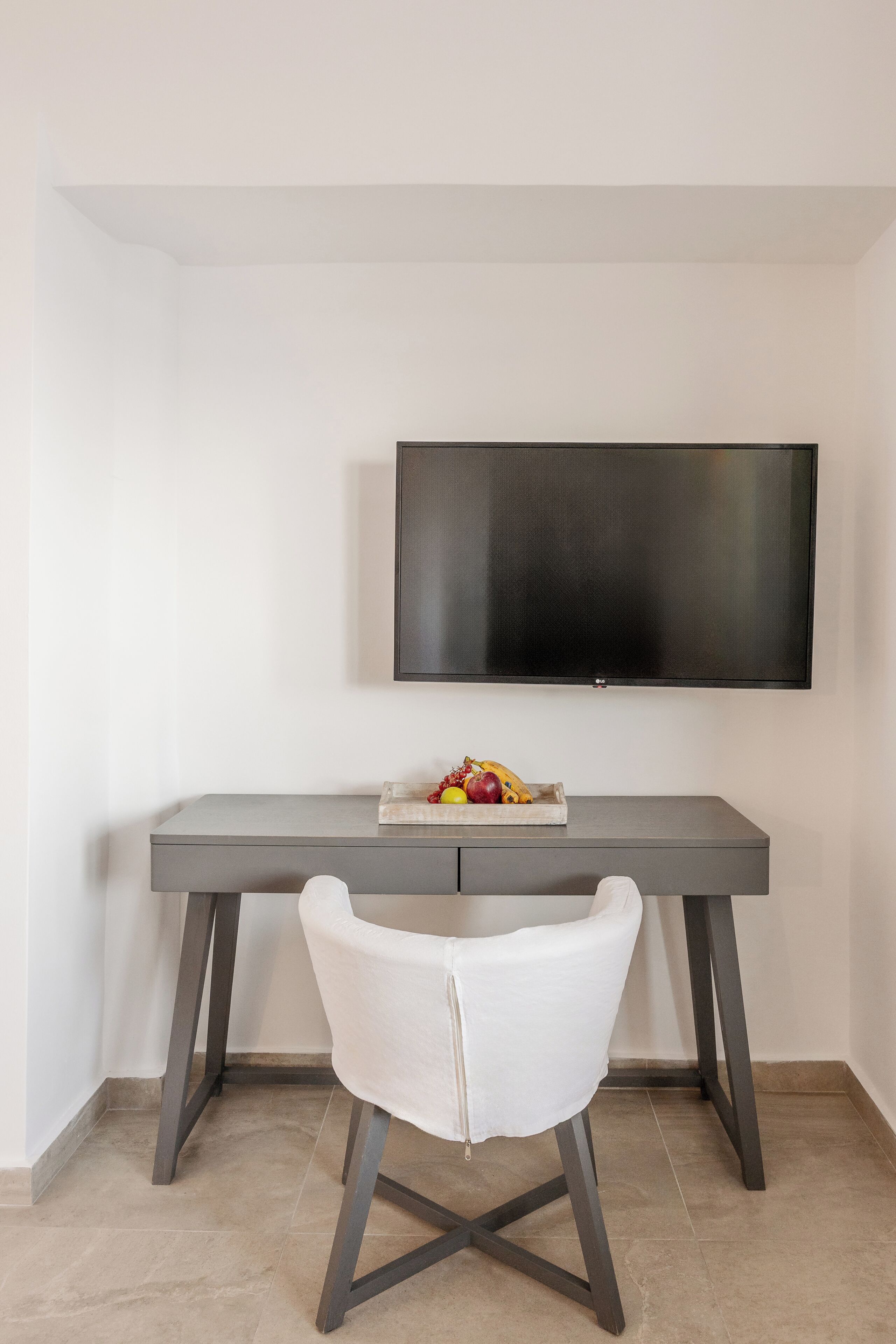 Quarto moderno com escrivaninha cinza, cadeira branca, televisão de tela plana e tigela de frutas na escrivaninha.