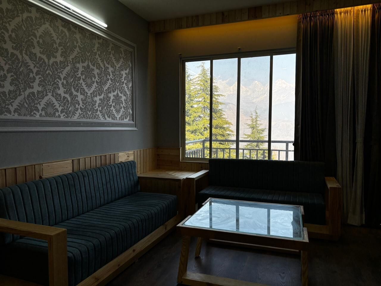 Quarto com sofá, mesa de centro de vidro e grande janela com vista para árvores e montanhas.