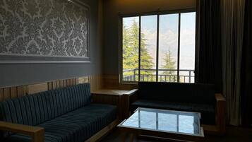 Quarto com sofá, mesa de centro de vidro e grande janela com vista para árvores e montanhas.
