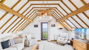 Quarto estilo loft com teto inclinado, vigas de madeira, um sofá, poltronas, uma TV e vista através de uma porta.