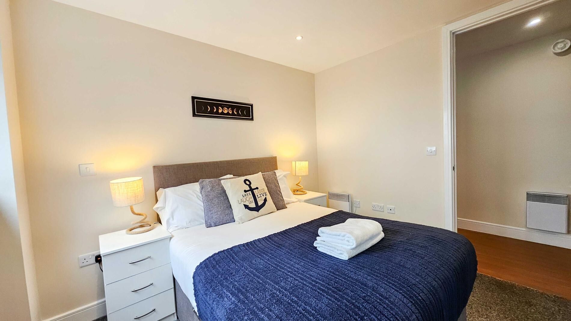 Quarto bem arrumado com cama, luminárias de cabeceira, mesa de cabeceira e relógio fixado na parede.
