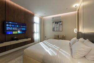 Quarto moderno com uma cama grande, TV fixada na parede exibindo serviços de streaming, ar condicionado de parede e decoração minimalista.