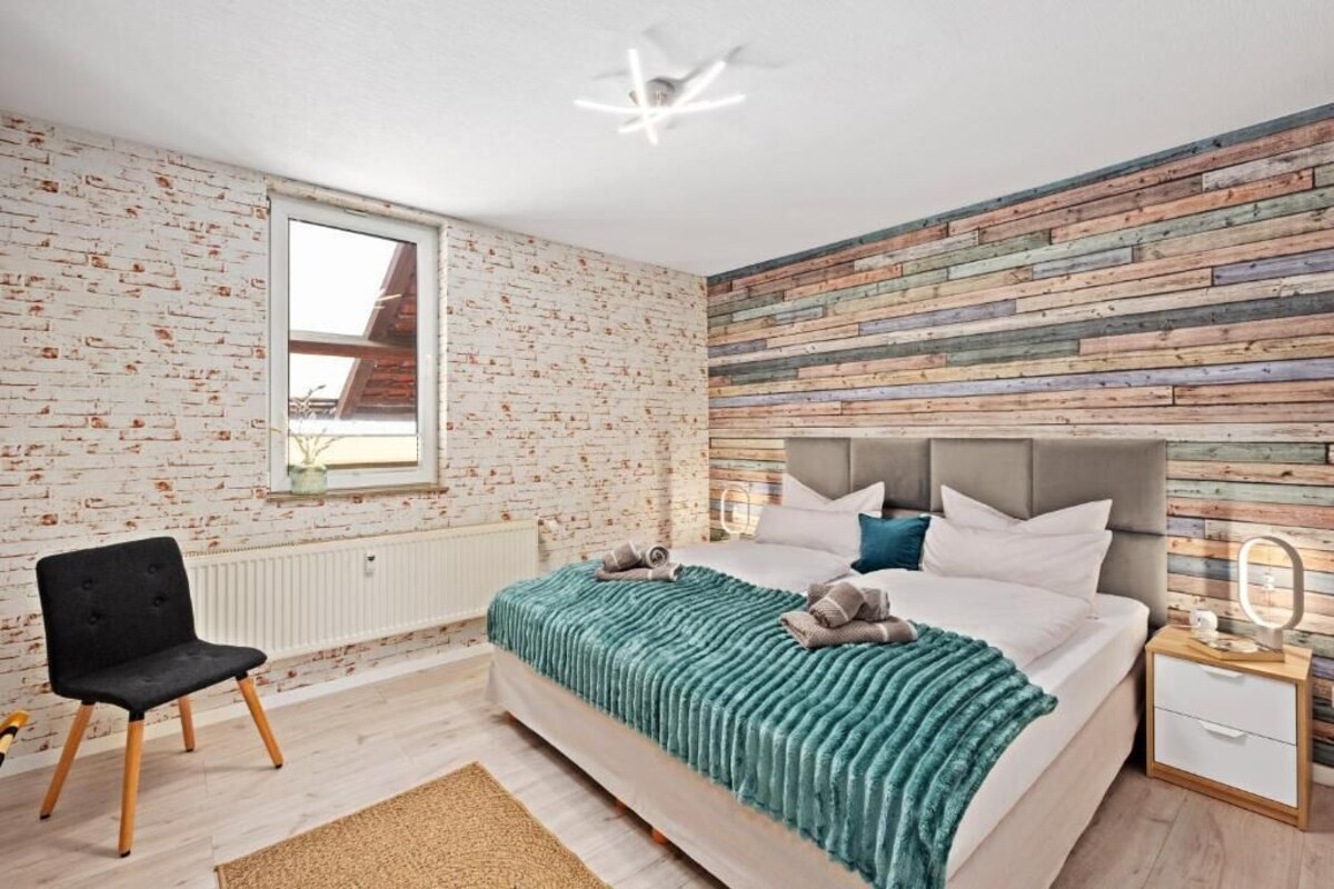 Quarto com parede em tijolos, cama com manta azul-turquesa, cadeira preta, mesa de cabeceira com espelho e janela com vista para um edifício.