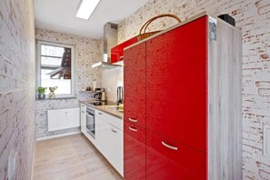 Cozinha com armários brancos, geladeira vermelha e parede de tijolos.