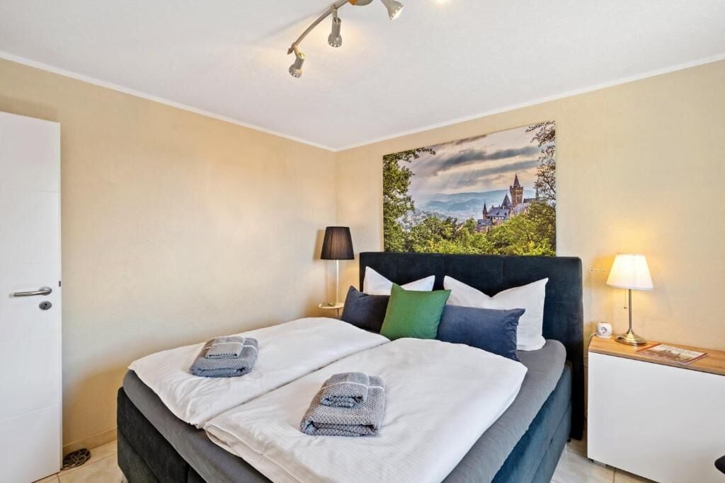Uma cama de casal com duas toalhas, cabeceira, abajures, mesa de cabeceira e uma pintura de paisagem urbana na parede.
