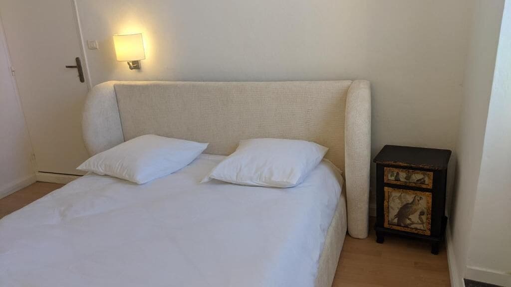 Uma cama com roupa de cama branca, uma mesa de cabeceira com abajur e uma pequena mesa com uma obra de arte.