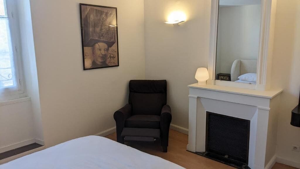 Quarto com cama, uma cadeira, lareira, luminária na parede e quadro emoldurado.