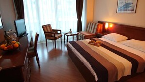 Quarto de hotel com cama, escrivaninha com computador, cadeira, mesinha de cabeceira com abajur e janela com cortinas.