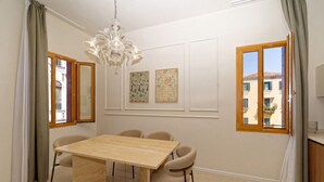 Uma sala de jantar com mesa e cadeiras de madeira, lustre, duas obras de arte floral emolduradas e janelas amplas com cortinas.