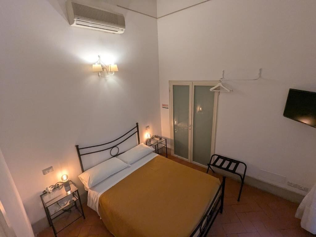 Quarto com cama, mesas de cabeceira, luminária pendente, TV fixada na parede e uma porta de vidro deslizante.