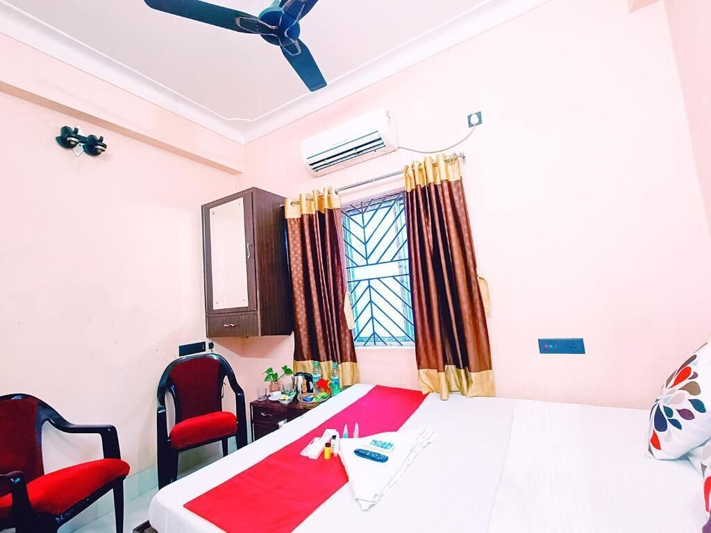 Quarto de hotel com cama, duas cadeiras vermelhas, ventilador de teto, ar condicionado, janela com cortinas e uma pequena mesa com objetos.