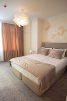 Quarto de hotel com uma cama grande, cabeceira, luminária fixada na parede, janela com cortinas e piso coberto por carpete.
