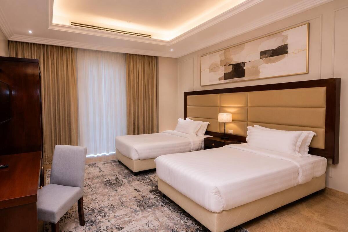 Quarto de hotel com duas camas, uma escrivaninha, uma cadeira, uma televisão e um quadro abstrato grande na parede.