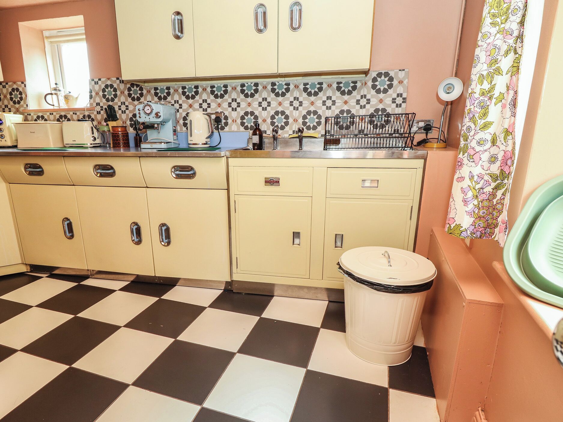 Uma cozinha com piso xadrez, armários cor de creme, backsplash ladrilhado e uma lixeira.