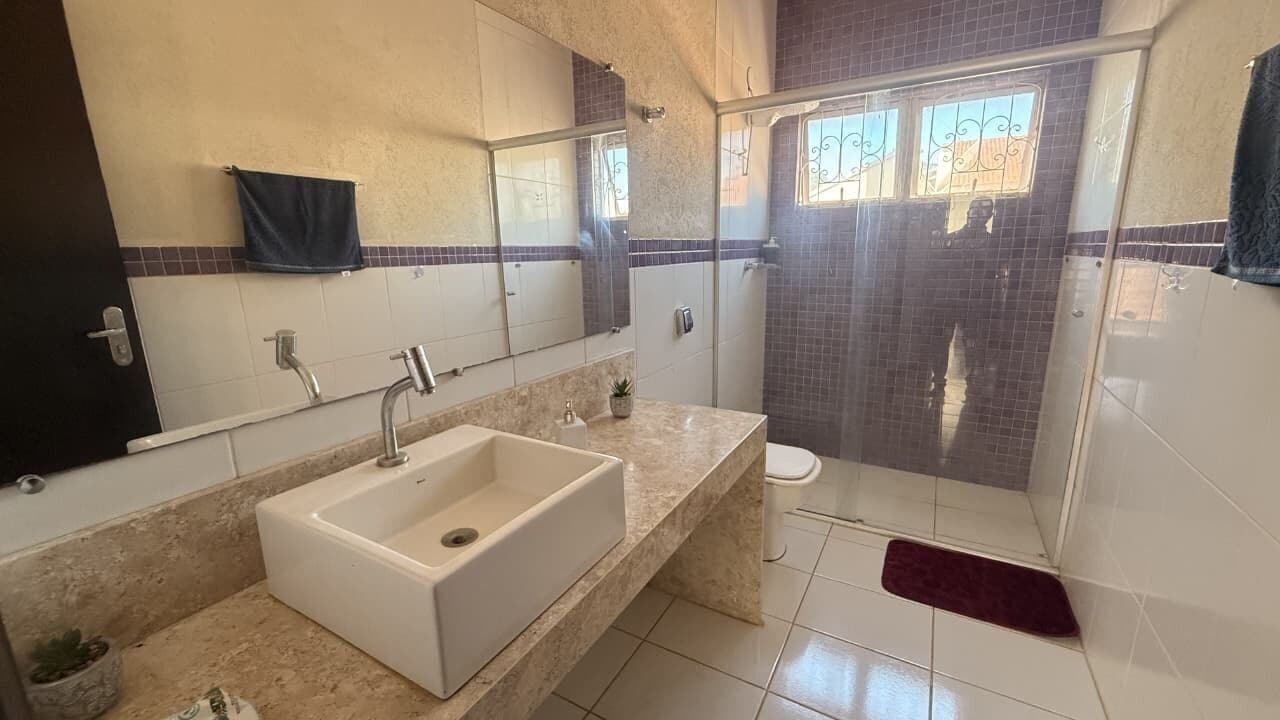 Casa de banho com uma pia branca, um espelho, chuveiro e um tapete de banho vermelho.