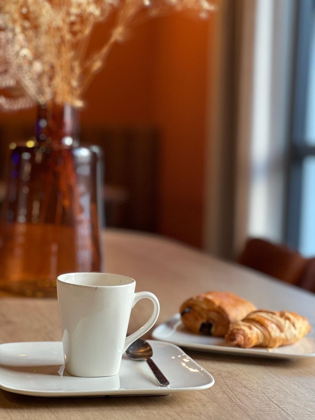 Uma mesa com uma xícara e pires brancos, uma colher e um prato com croissants.