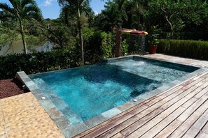Uma piscina retangular com um deck de madeira ao redor, situada num jardim exuberante com árvores e céu claro.