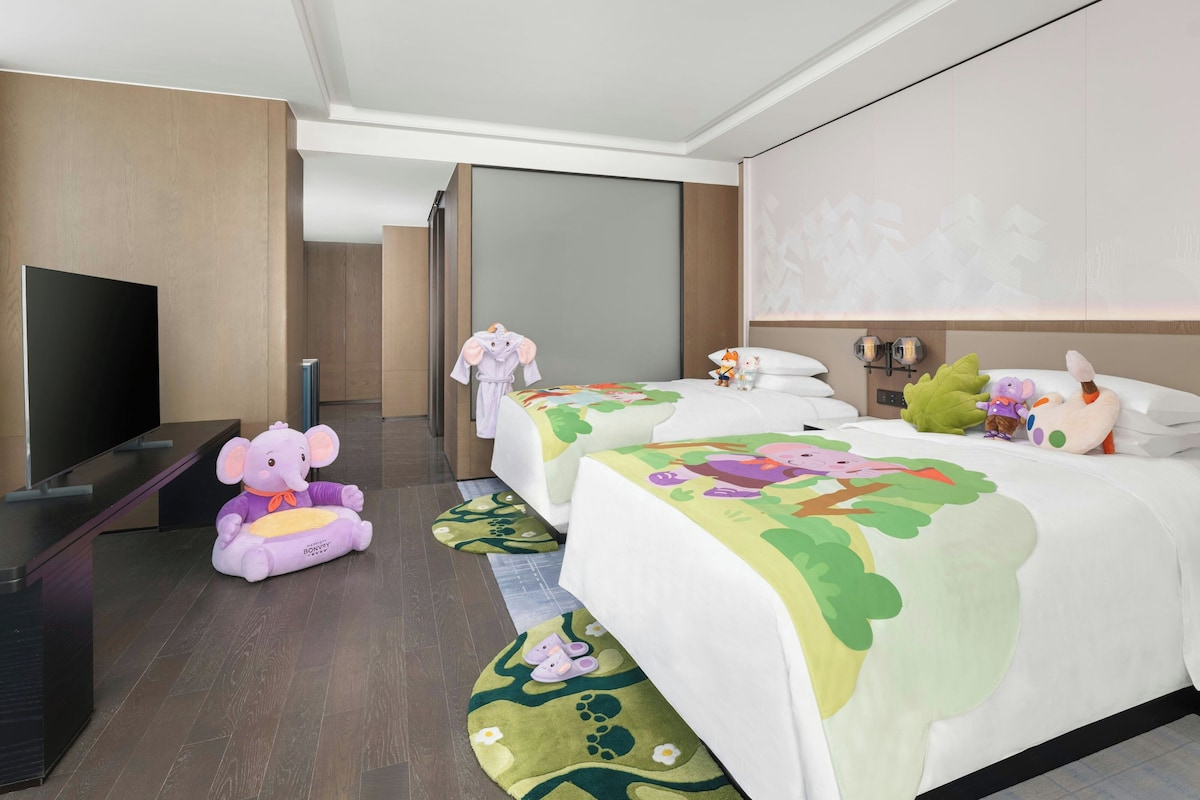 Quarto de hotel com televisão de tela plana grande, duas camas com roupa de cama colorida, brinquedos de pelúcia e design moderno.