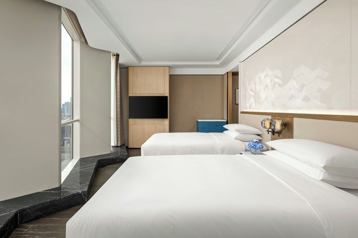 Quarto de hotel com duas camas, televisão de tela plana, mesa de cabeceira com um vaso de flores e vista para a cidade.