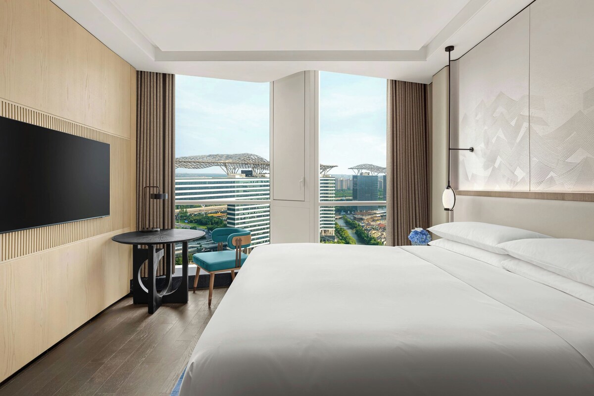 Quarto de hotel com cama grande, televisão de tela plana, mesa redonda pequena com uma cadeira e vista panorâmica da cidade através de amplas janelas.