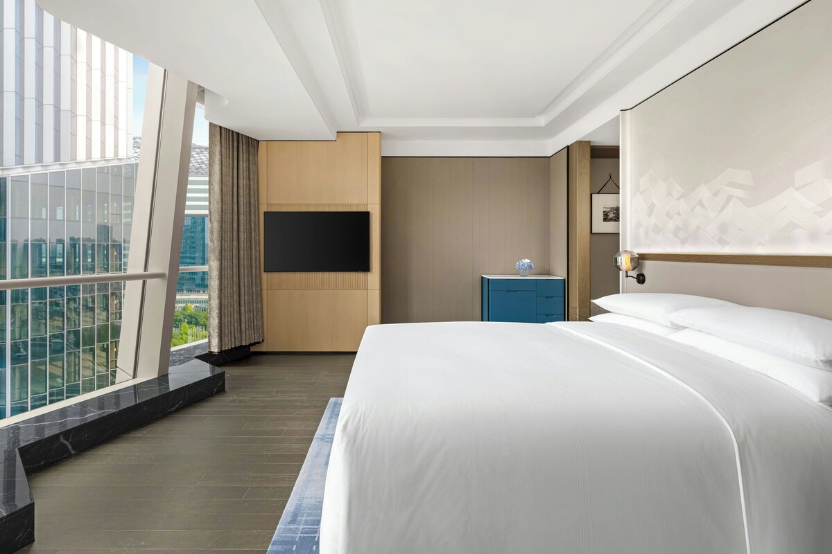 Quarto de hotel com uma cama grande, televisão de tela plana, uma escrivaninha pequena e vista para o horizonte urbano através de janelas do chão ao teto.