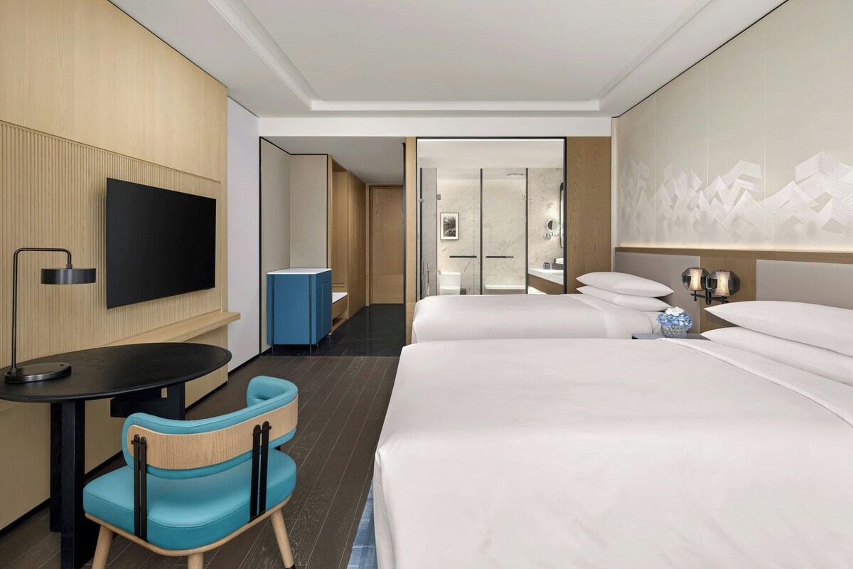 Quarto de hotel com cama, escrivaninha, cadeira, televisão e banheiro com chuveiro e vaso sanitário.