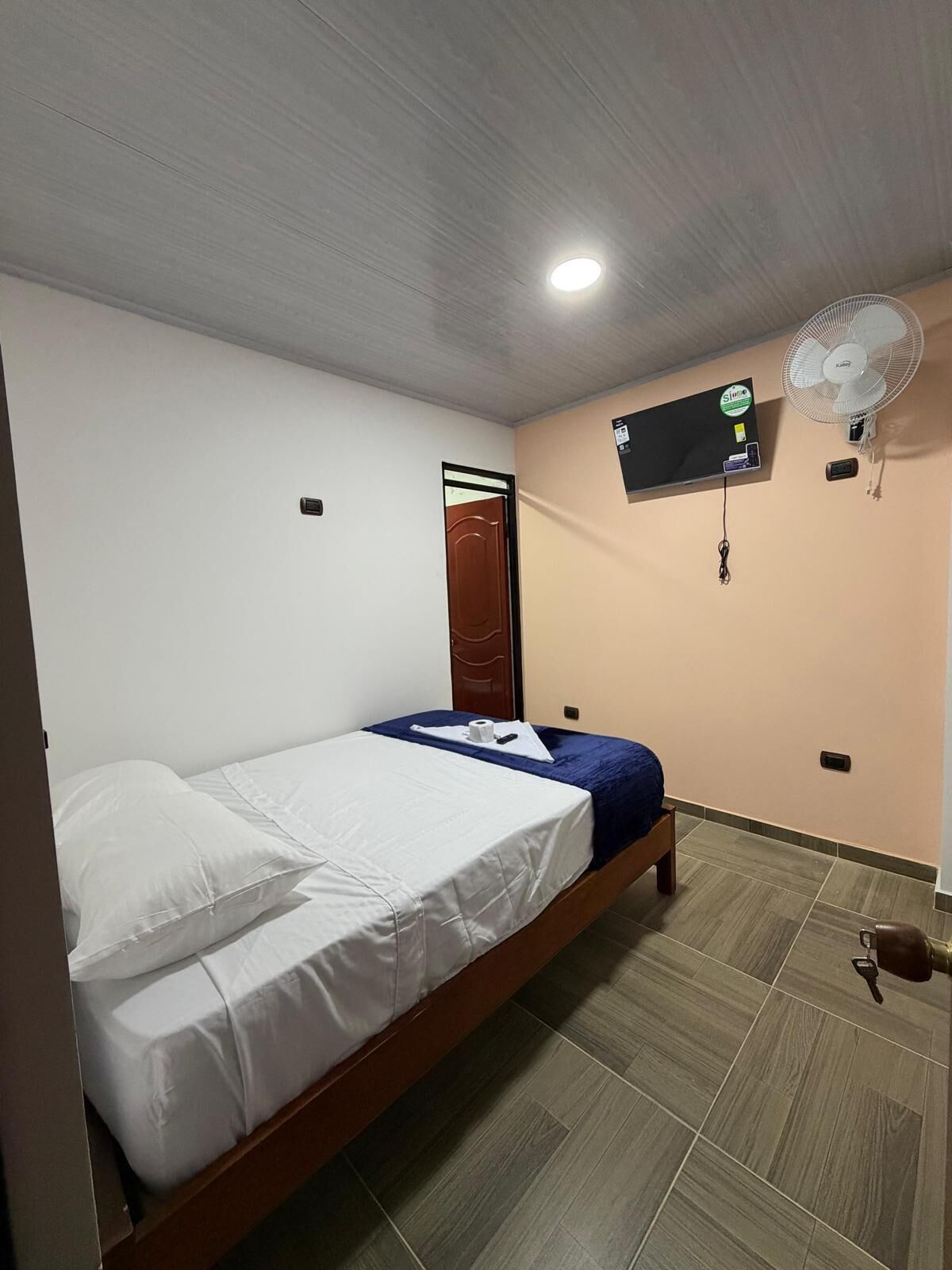 Uma cama de solteiro com roupa de cama branca, cabeceira de madeira, televisão montada na parede, ventilador de teto e uma porta com moldura de madeira.