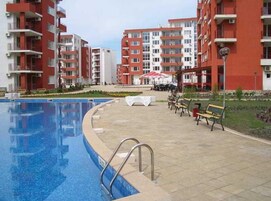 Piscina com escada de metal, calçada e edifícios de apartamentos vermelhos e brancos ao fundo.