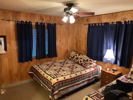 Quarto com parede revestida em madeira, cama com colcha estampada, mesa de cabeceira com abajur e ventilador de teto.