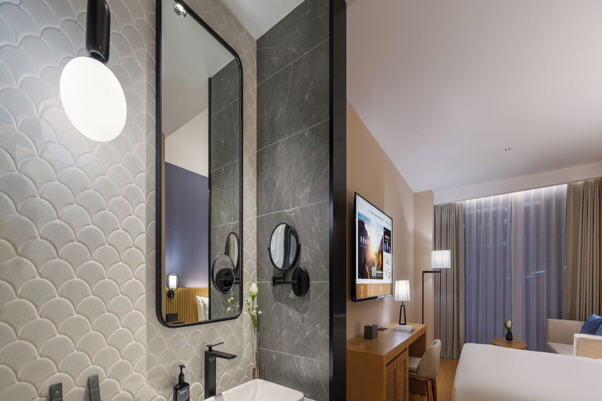 Quarto de hotel com um espelho grande, pia, cama, escrivaninha e televisão.