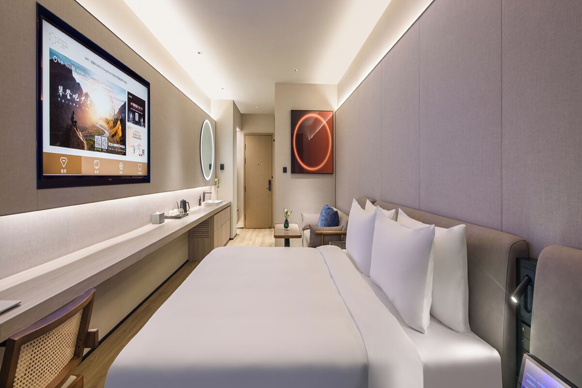 Quarto de hotel moderno com uma cama grande, uma escrivaninha, uma televisão e um candeeiro circular.