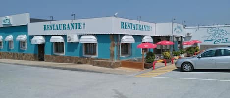 Um edifício com uma placa que diz 'RESTAURANTE' e 'HOTEL'. Há mesas e cadeiras ao ar livre, e um carro estacionado na frente.