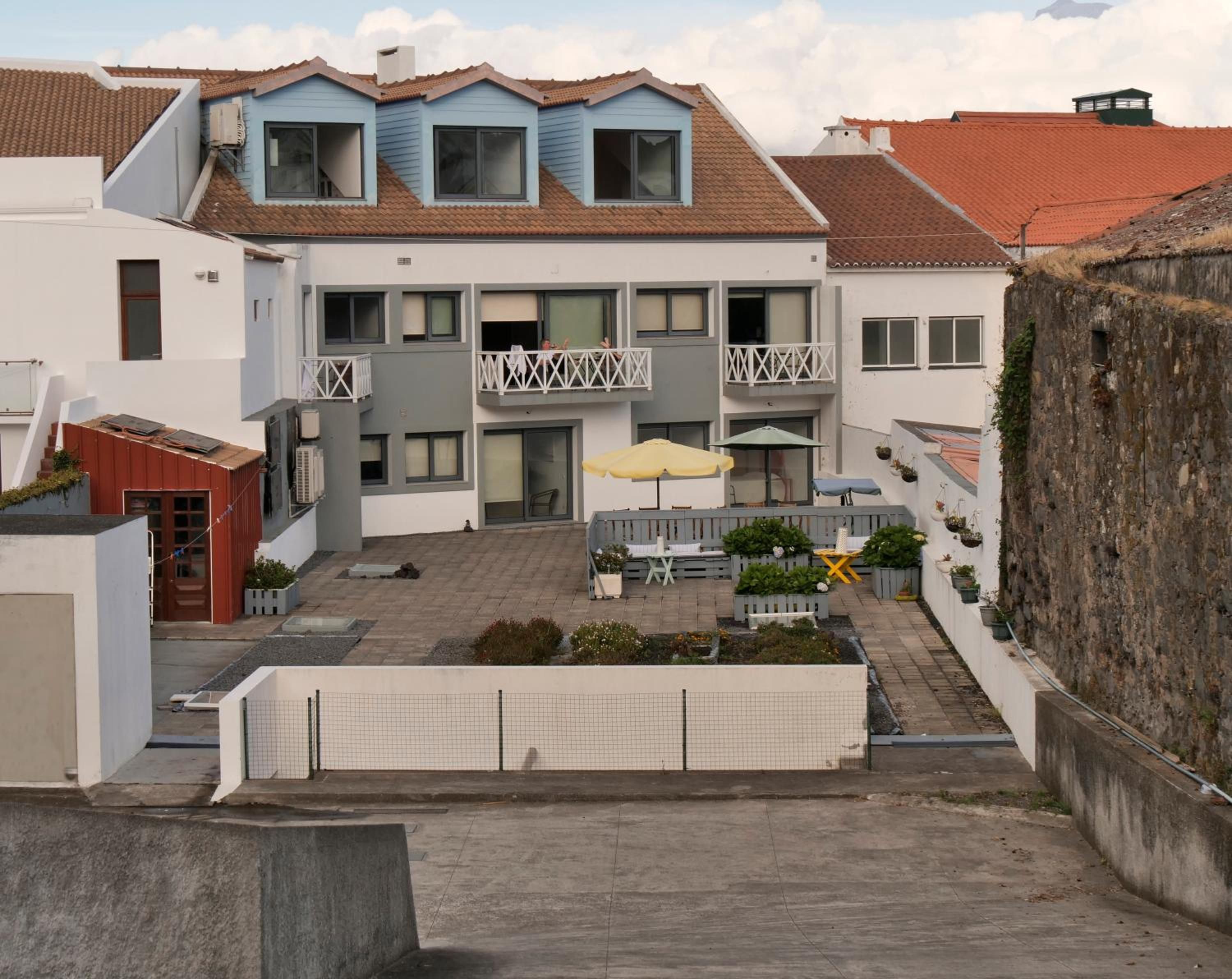 Edifício residencial com vários andares, varandas, um pátio com mobiliário exterior e uma parede de pedra.