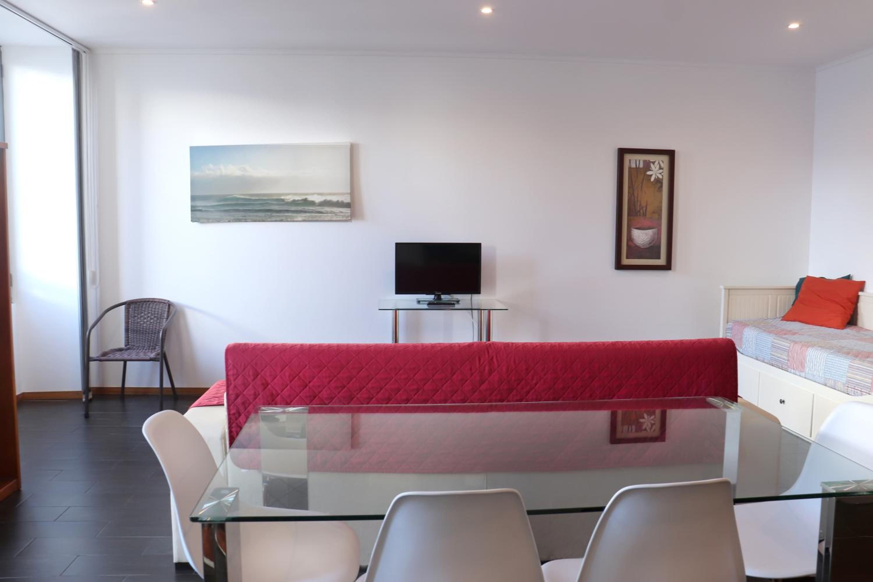 Quarto moderno com mesa de jantar de vidro, cadeiras brancas, cama com almofadas vermelhas, televisão sobre um suporte e obras de arte nas paredes.