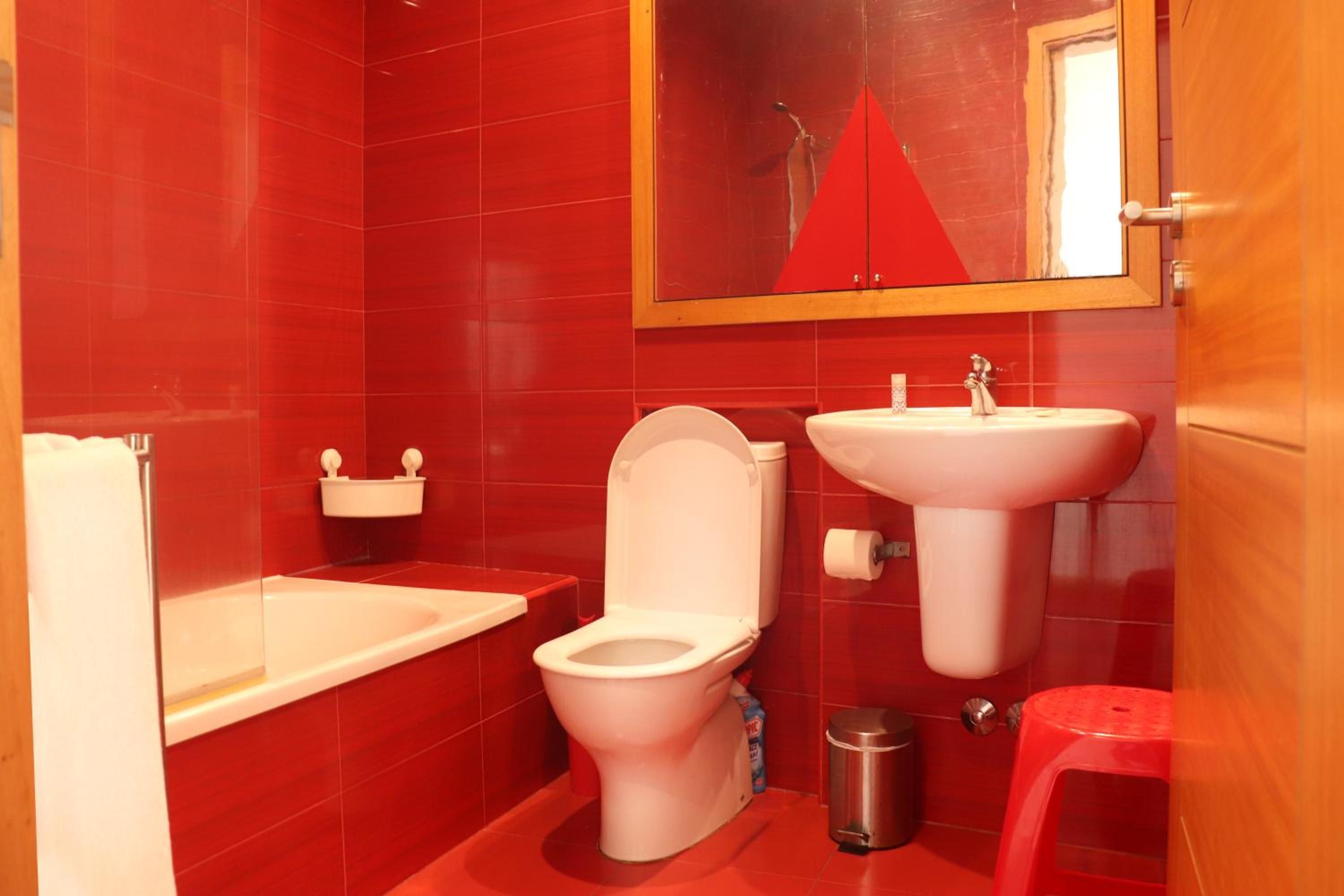 Casa de banho com azulejos vermelhos, sanita branca, lavatório branco, banheira e espelho sobre o lavatório.