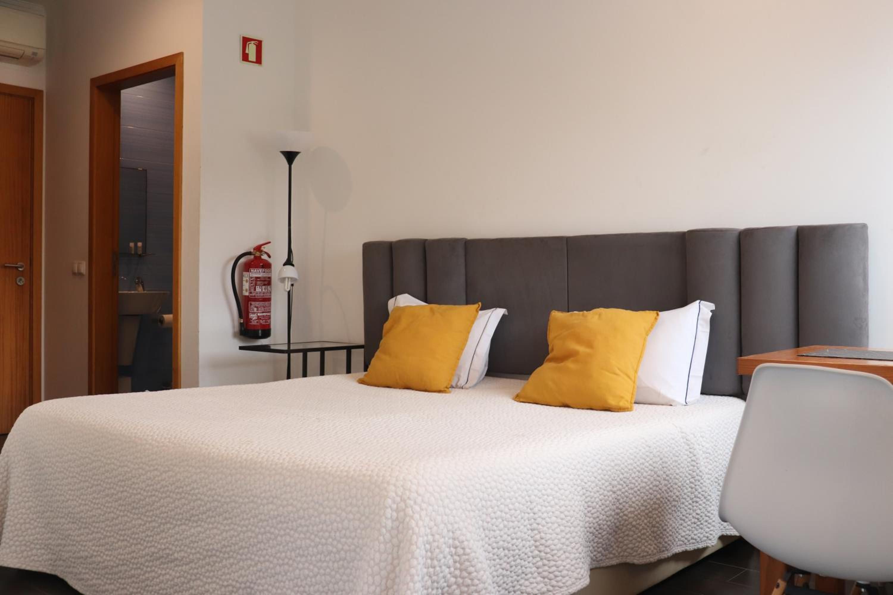 Quarto de hotel com cama, mesa de cabeceira, cadeira, extintor de incêndio e porta.